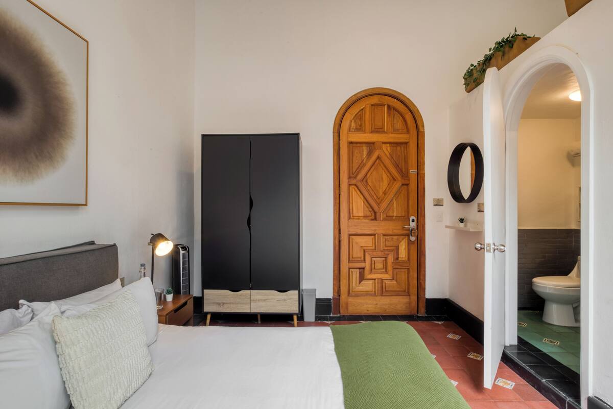 Quarto com cama, guarda-roupa, porta e banheiro visível por uma porta aberta.