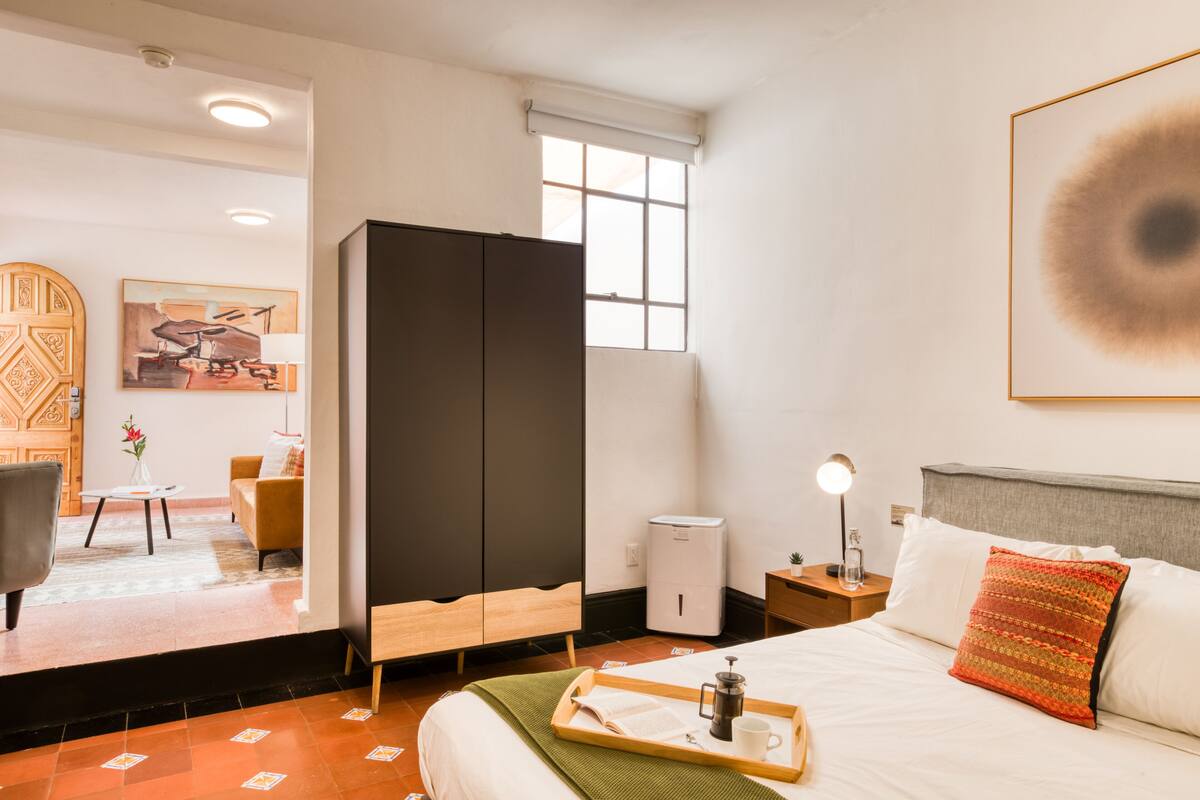 Quarto de hotel com cama, mesa de cabeceira, abajur, guarda-roupa, janela e um quadro na parede.