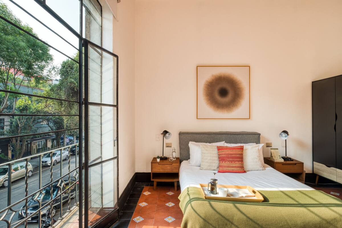 Um quarto com uma janela grande, uma cama com manta verde, duas mesas de cabeceira com luminárias e um quadro na parede.