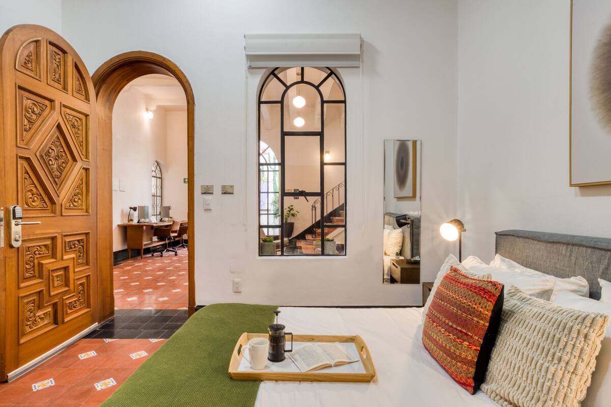 Quarto de hotel com uma cama, um cobertor verde, uma bandeja com um livro e uma caneca, e uma janela com vista para o exterior.