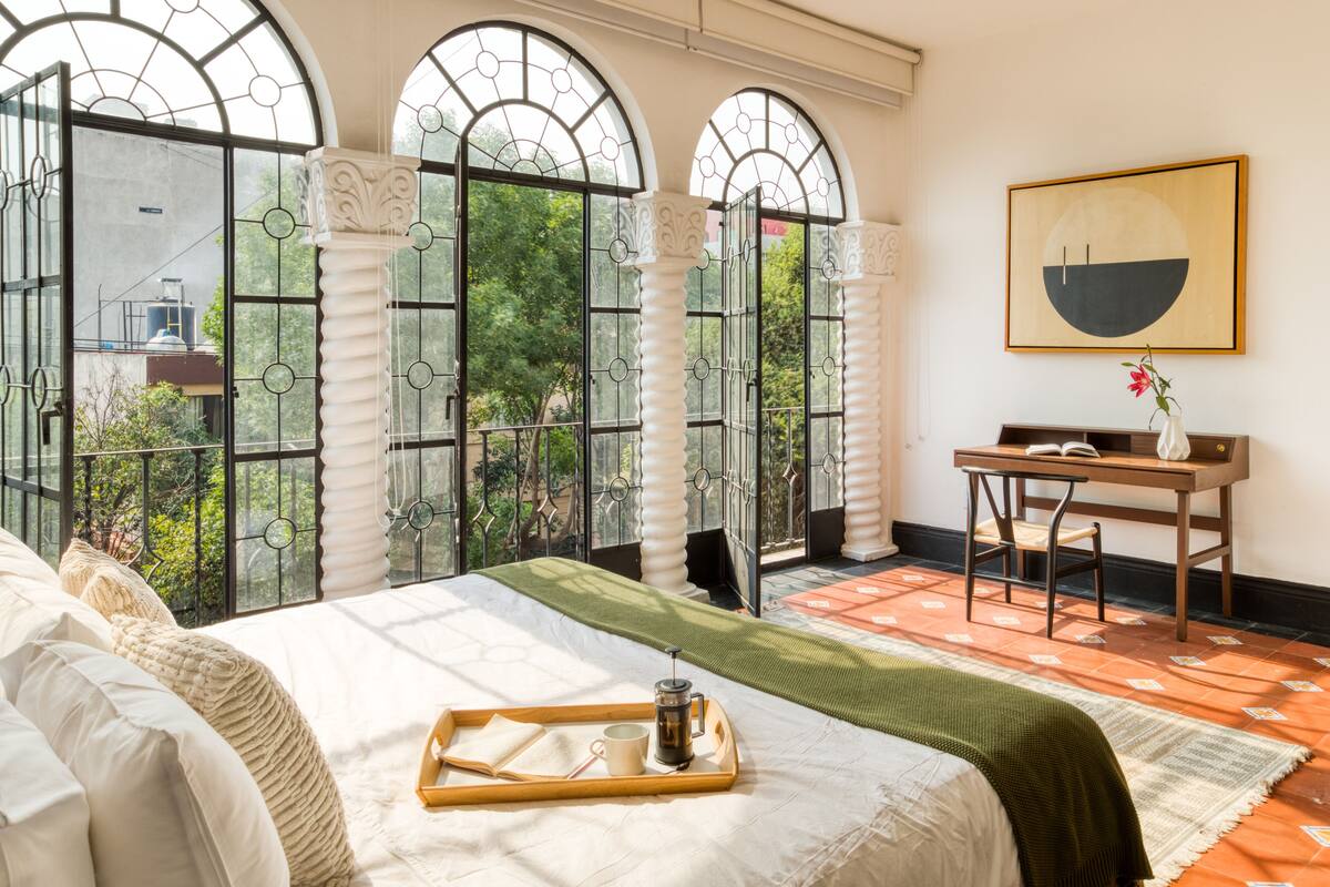 Quarto com uma cama grande, mesa de cabeceira de madeira, uma cadeira e vista através de janelas amplas.