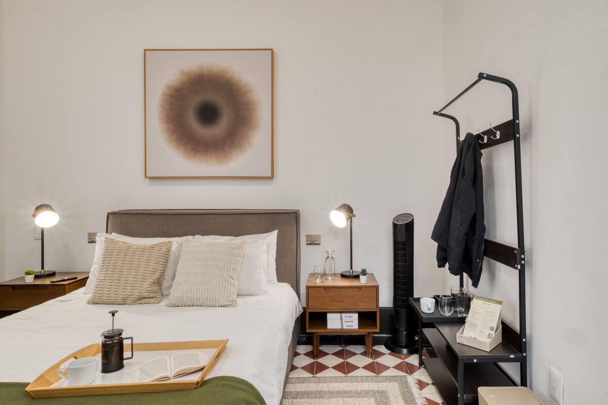 Quarto de hotel bem organizado com cama, mesas de cabeceira, máquina de café, bandeja com xícara e livro, e um cabideiro.