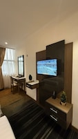 Quarto de hotel com televisão de tela plana na parede, escrivaninha com cadeira, uma pequena planta e um armário de madeira.