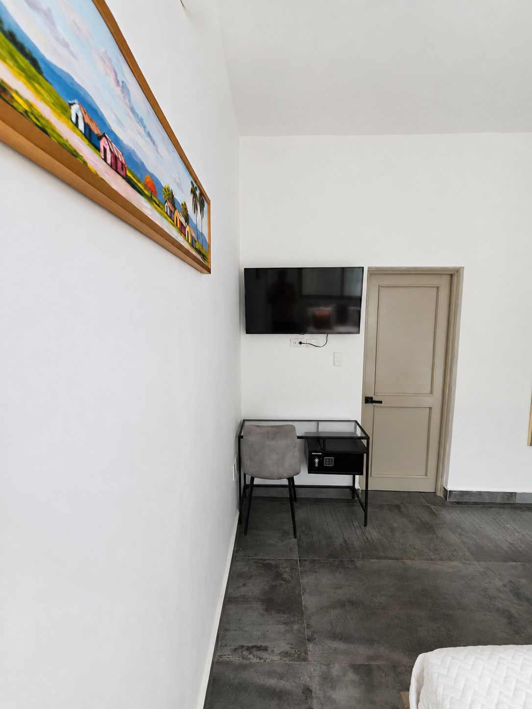 Quarto com cama, escrivaninha com cadeira, televisão fixada parede e uma porta.