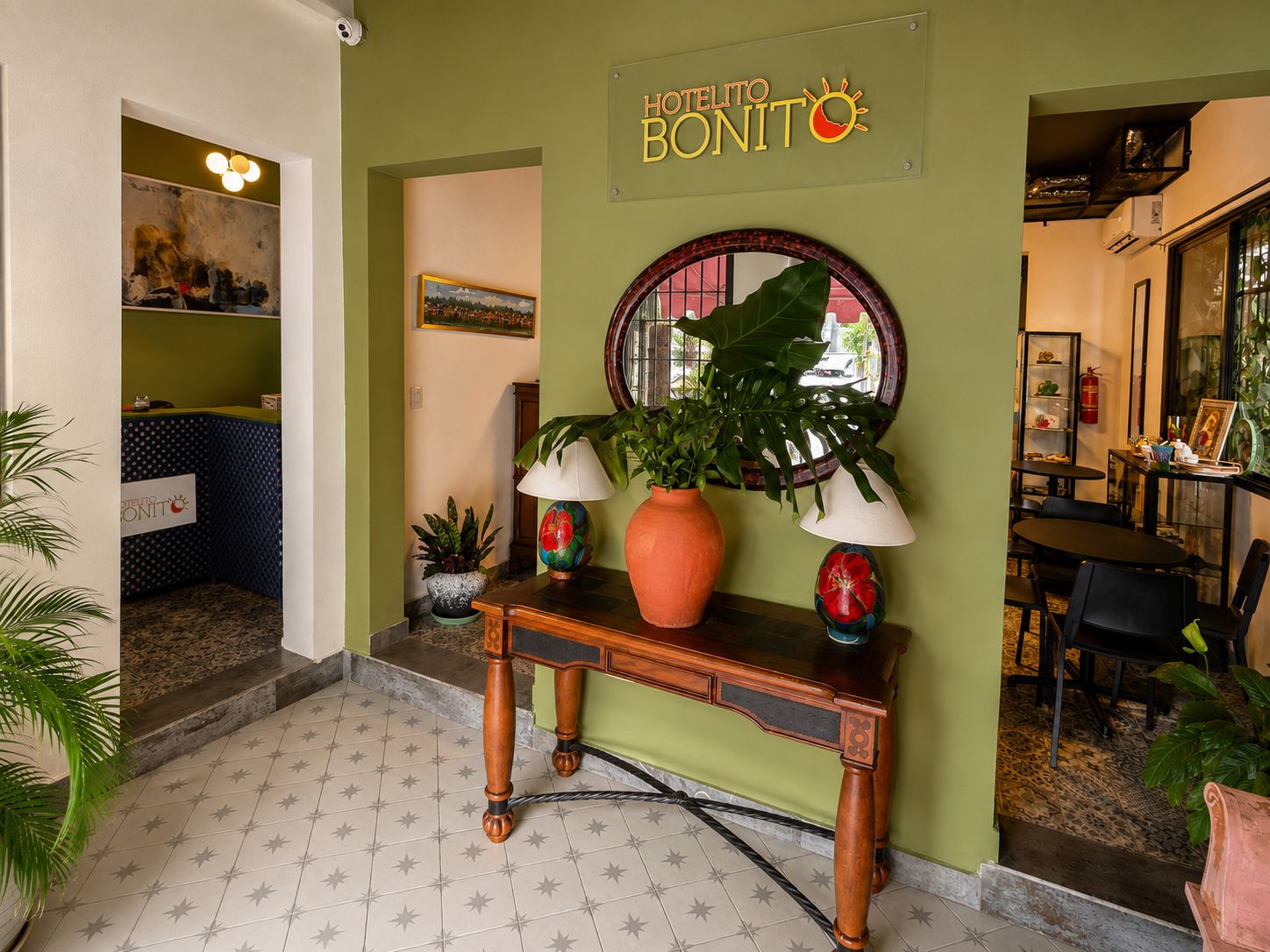 Um saguão de hotel com balcão de recepção, uma grande planta em vaso, uma placa na parede e uma área de refeições com cadeiras e mesas.