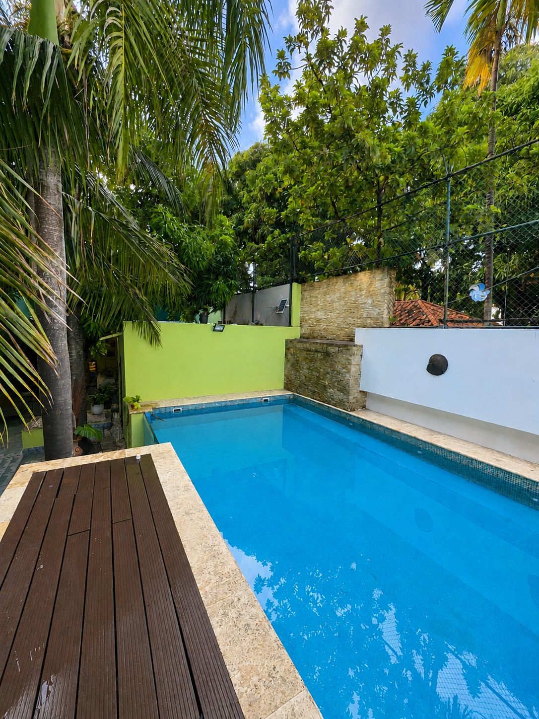 Piscina com deck de madeira, rodeada por vegetação exuberante e um muro de pedra.