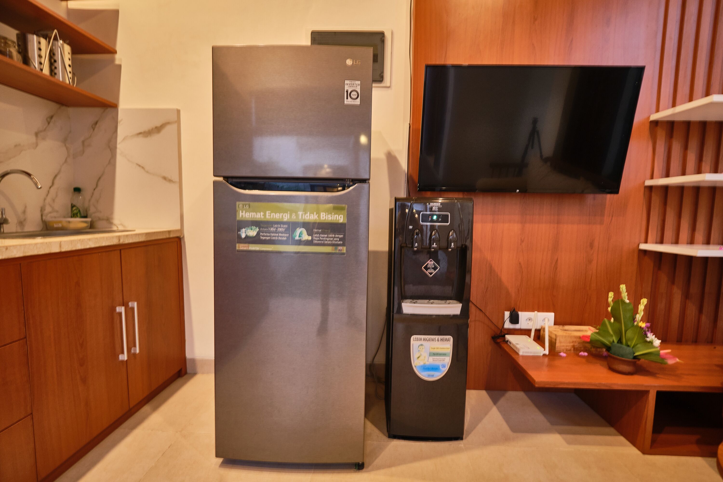 Uma cozinha com geladeira de aço inoxidável, bebedouro, armários de madeira, painel de backsplash de mármore e uma TV de tela plana na parede.
