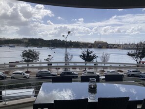 Mesa com cadeiras e um vaso de vidro, com vista para um porto com barcos e um edifício ao longe.