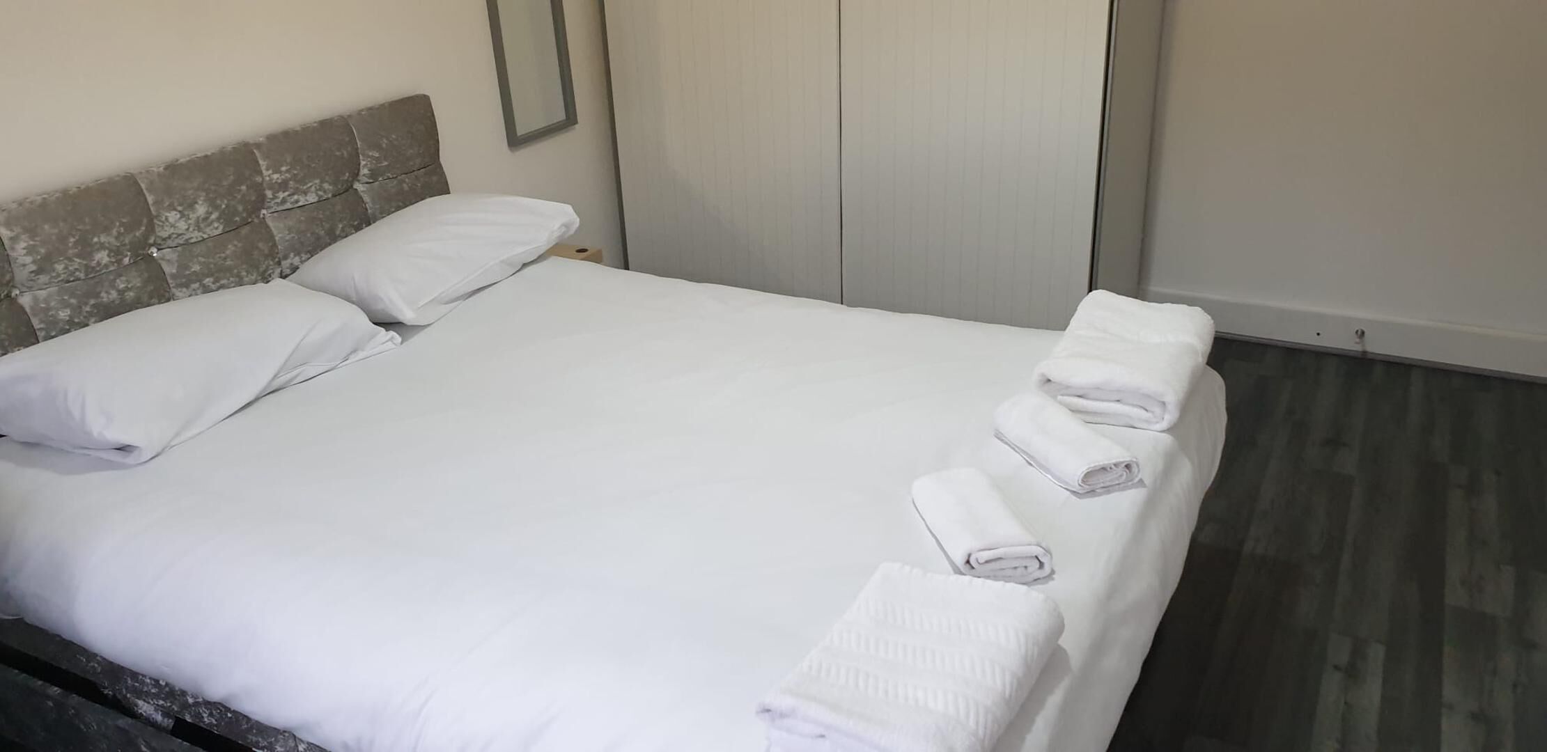 Cama bem arrumada com lençóis e almofadas brancos, cabeceira de madeira e um conjunto de toalhas ao lado.