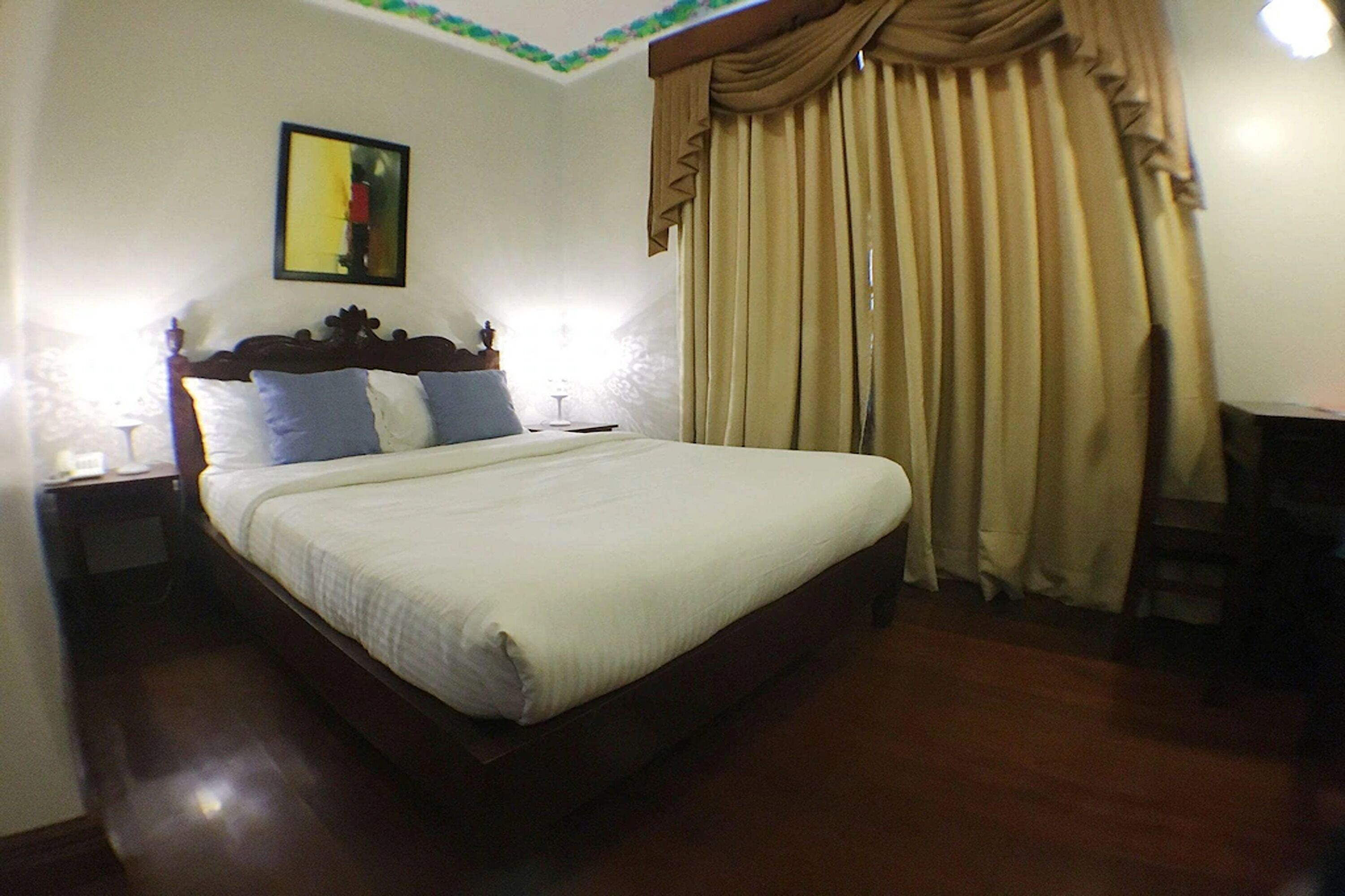 Uma cama bem arrumada com cabeceira de madeira, uma mesa de cabeceira com abajur e uma janela grande com cortinas bege.