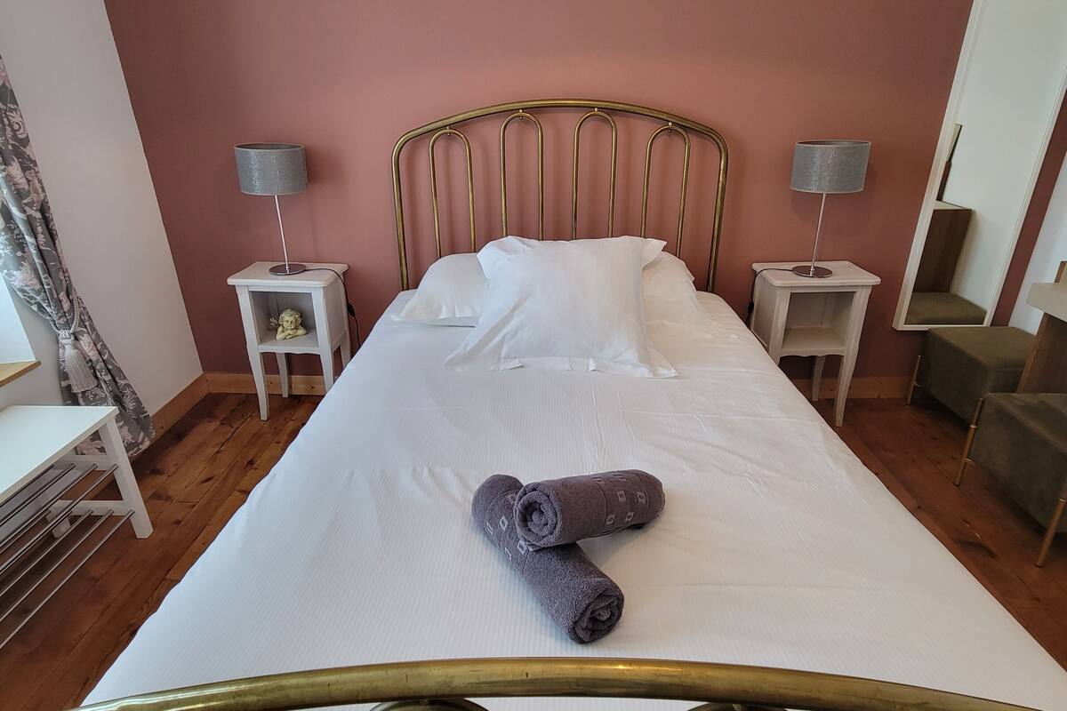 Cama bem arrumada com lençóis brancos, duas mesas de cabeceira com abajures, uma toalha dobrada sobre a cama e um espelho na parede.