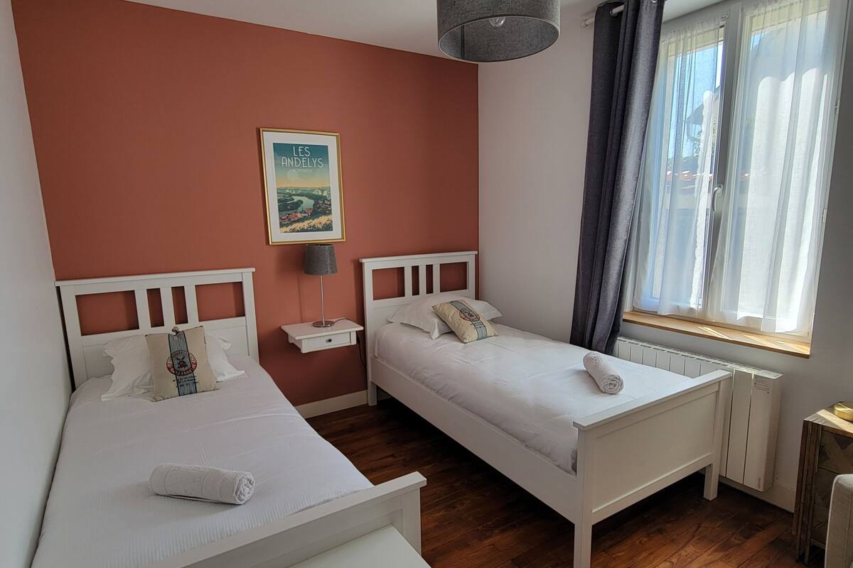 Quarto com duas camas de solteiro, uma pequena mesa de cabeceira com abajur, radiador e janela com cortinas.