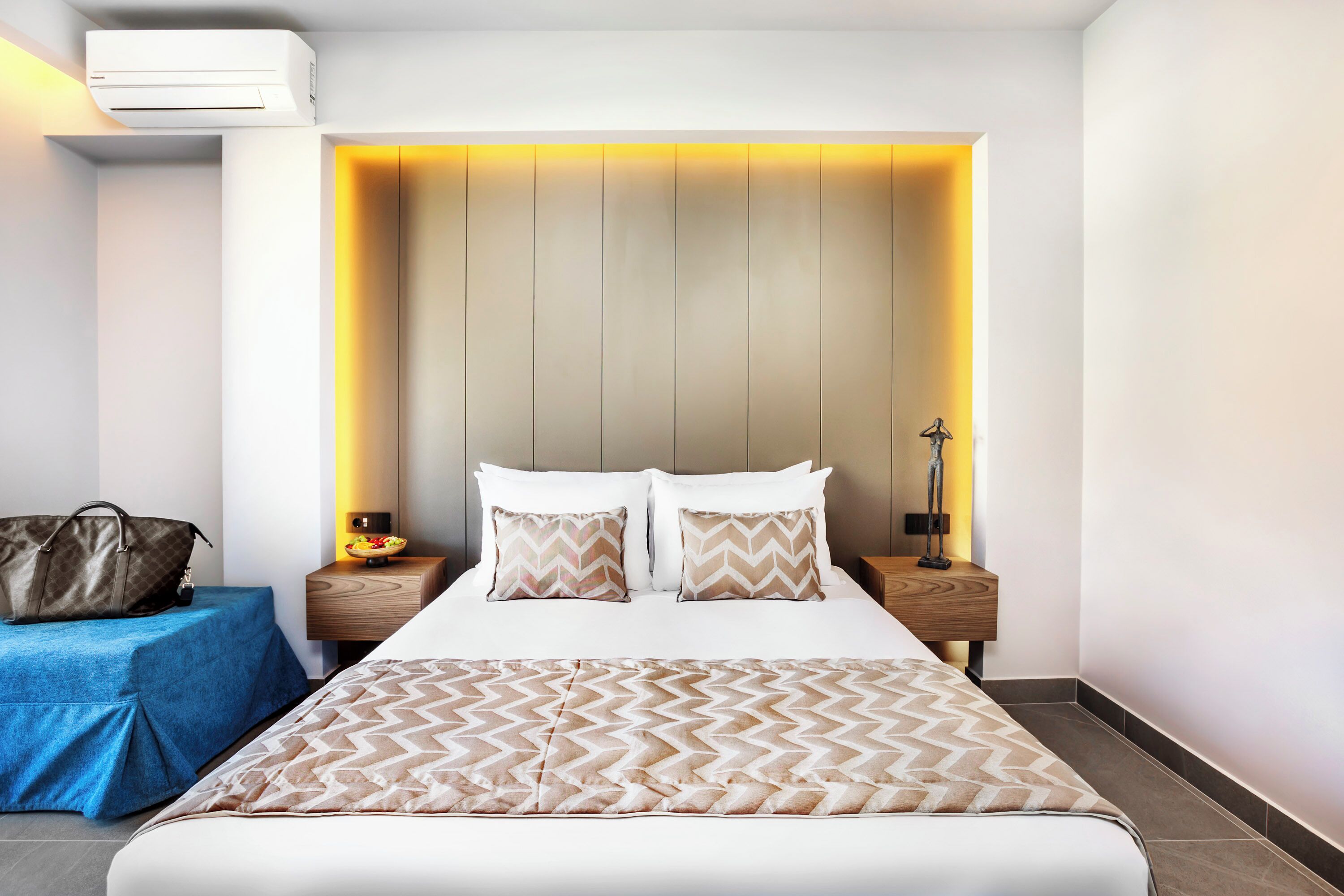 Quarto de hotel moderno com uma cama grande, uma mesa de cabeceira com abajur, um criado-mudo e uma cadeira azul.