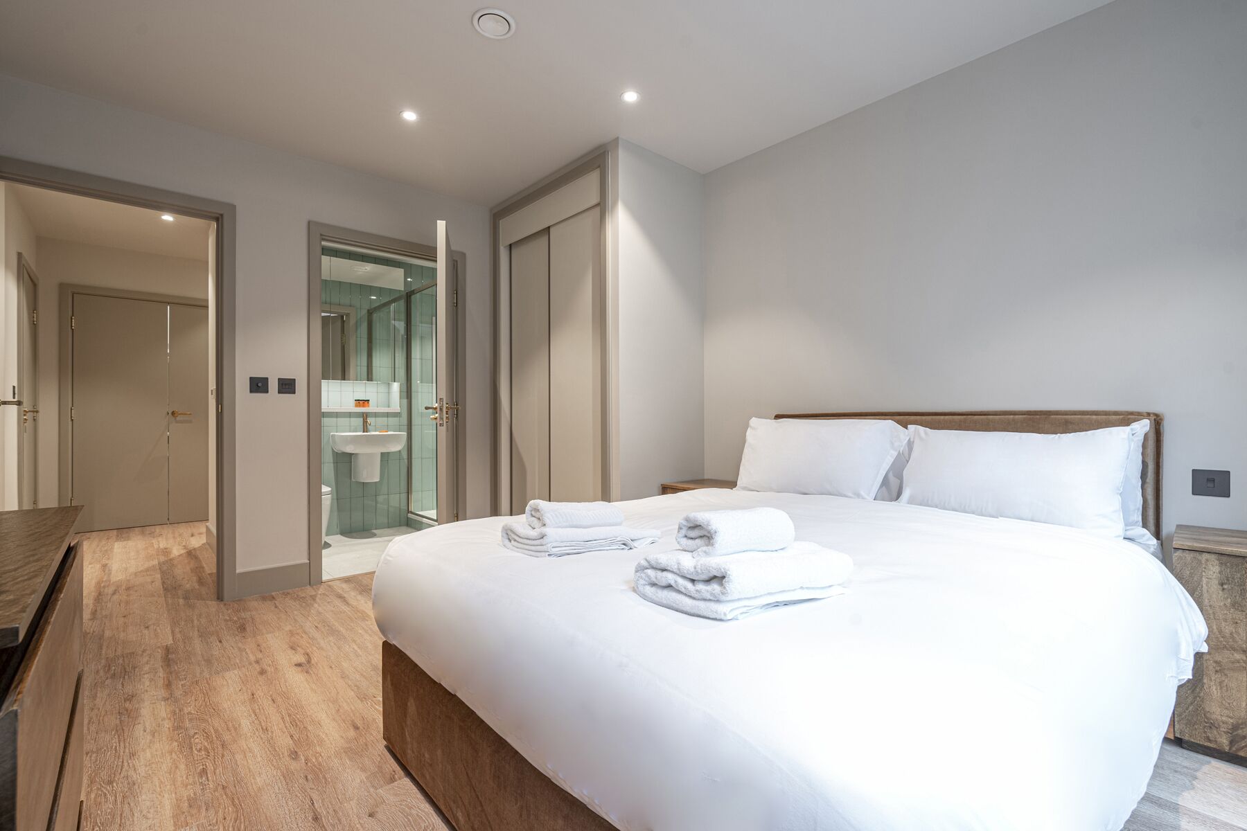 Quarto de hotel moderno com uma cama grande, um banco de madeira, chuveiro em cabine de vidro e uma pequena mesa de cabeceira.