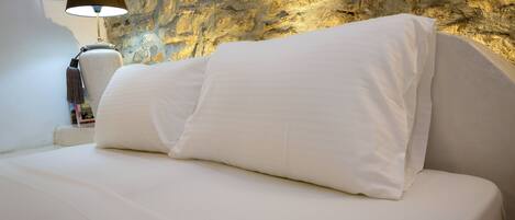 Cama com roupa de cama branca, luminária com cúpula marrom e parede de pedra ao fundo.