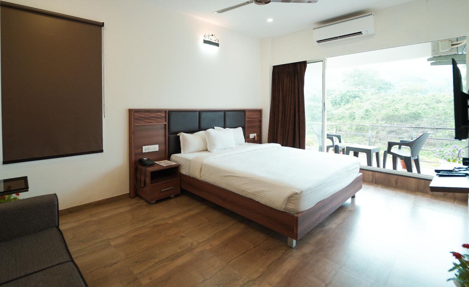 Quarto de hotel com uma cama grande, mesa de cabeceira, escrivaninha com cadeiras e vista para o verde lá fora.