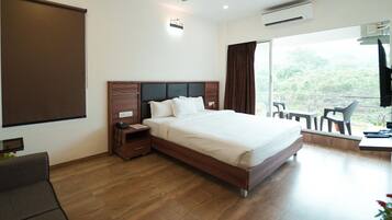 Quarto de hotel com uma cama grande, mesa de cabeceira, escrivaninha com cadeiras e vista para o verde lá fora.