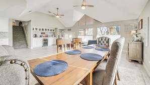Uma sala ampla com uma mesa de jantar de madeira, várias cadeiras, ventiladores de teto e uma cozinha ao fundo.