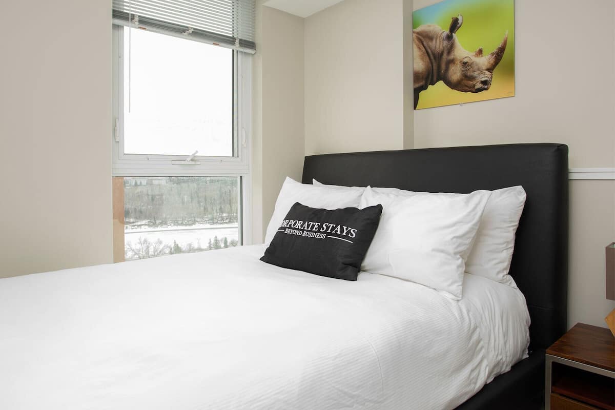 Quarto de hotel com cama, janela com persianas, pintura de um rinoceronte e travesseiro com texto.