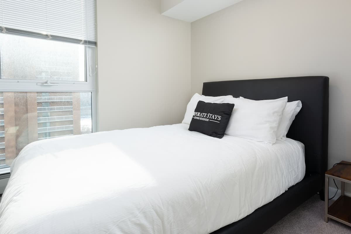 Cama bem arrumada com lençóis brancos e almofada preta com texto, uma janela com persianas e uma parede lisa.