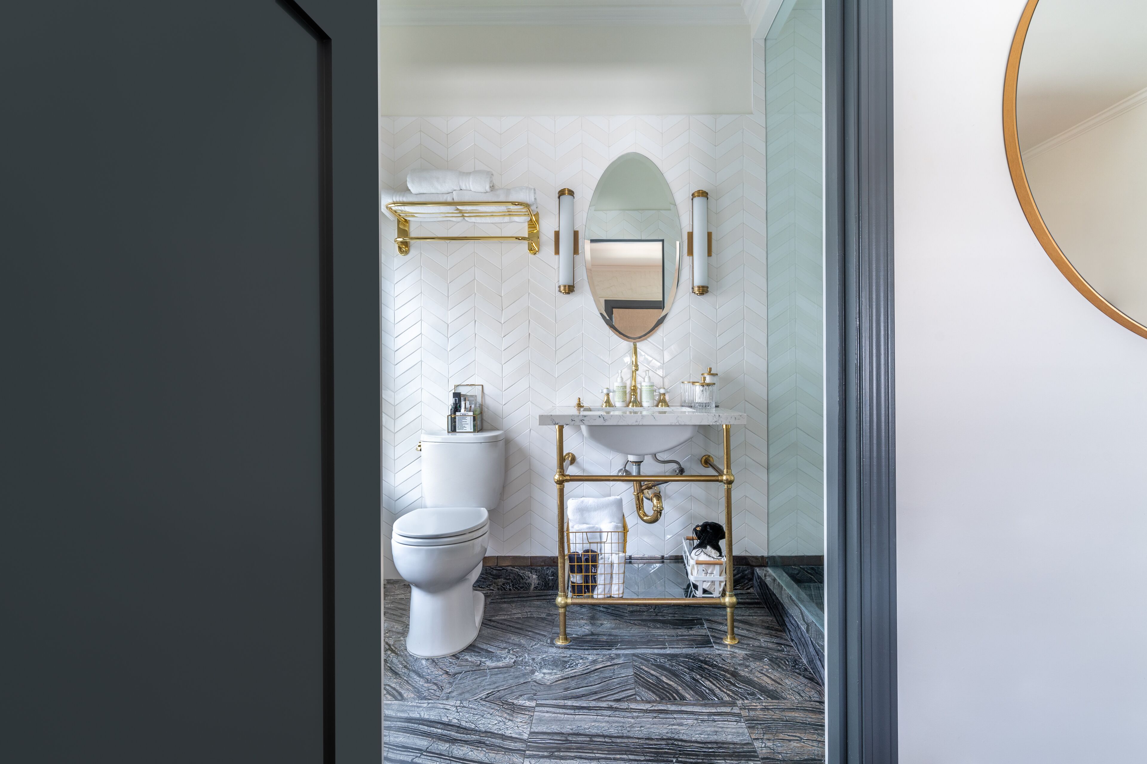Casa de banho com sanita branca, toucador dourado com espelho e chão com padrão herringbone.