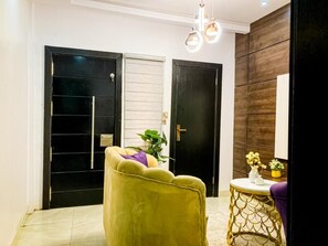 Um quarto com uma poltrona amarela, uma mesa redonda com base dourada e duas portas pretas com um painel branco no centro.