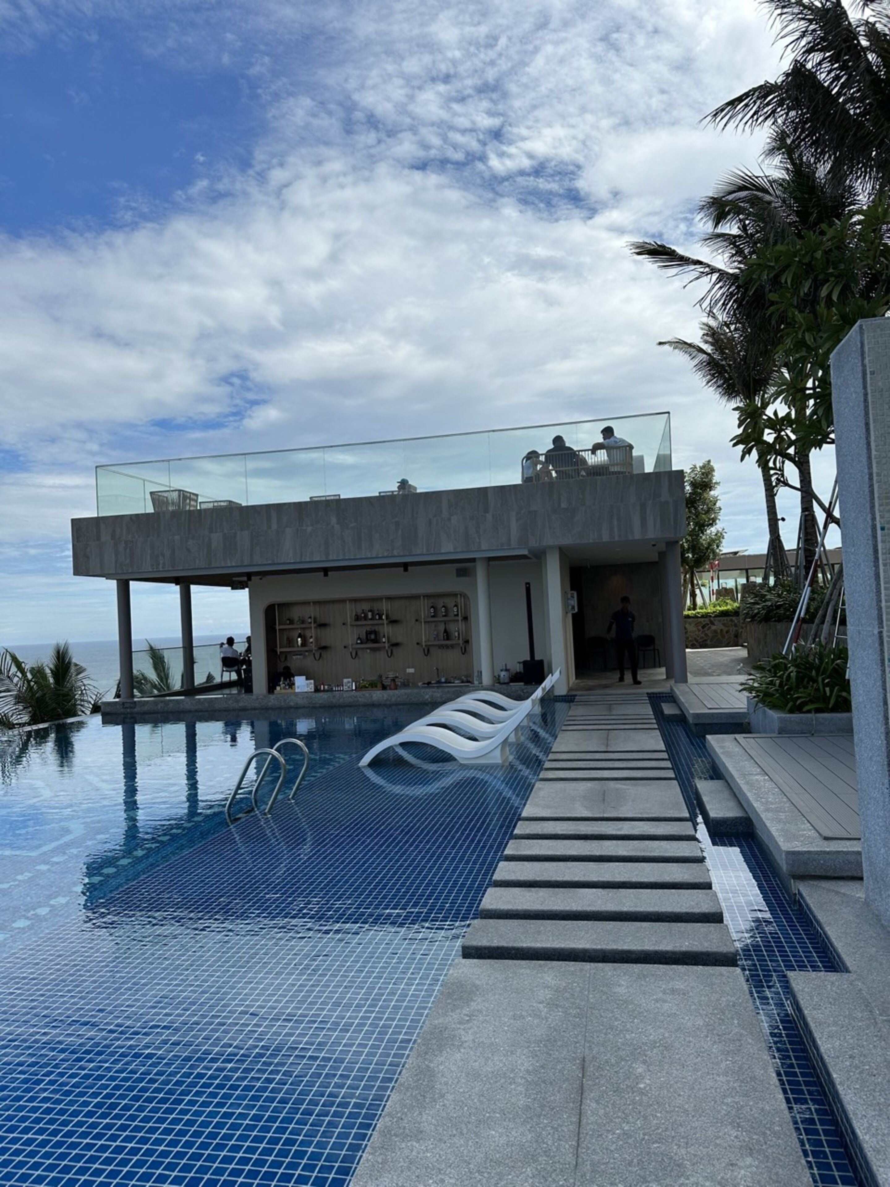 Uma área de piscina moderna com bar e lounge com paredes de vidro, cercada por palmeiras e vista desobstruída do oceano.