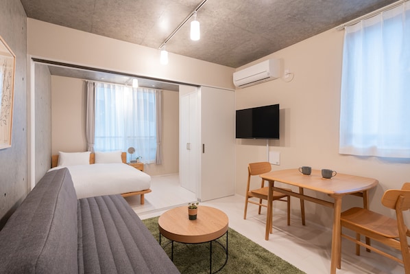 Quarto de hotel moderno com cama, sofá, mesa de jantar com cadeiras, uma mesinha redonda com planta e TV fixada na parede.