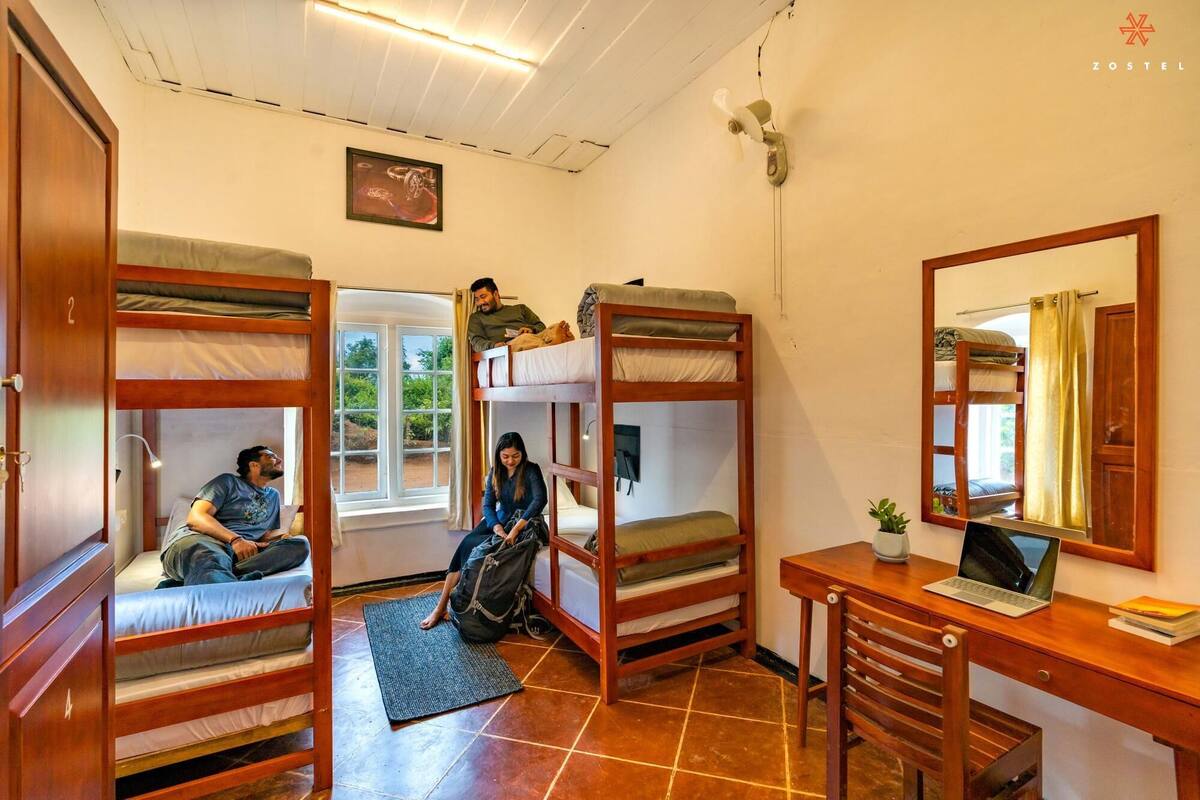 Quarto em dormitório com beliches, uma escrivaninha com um laptop e uma janela com vista para o verde.