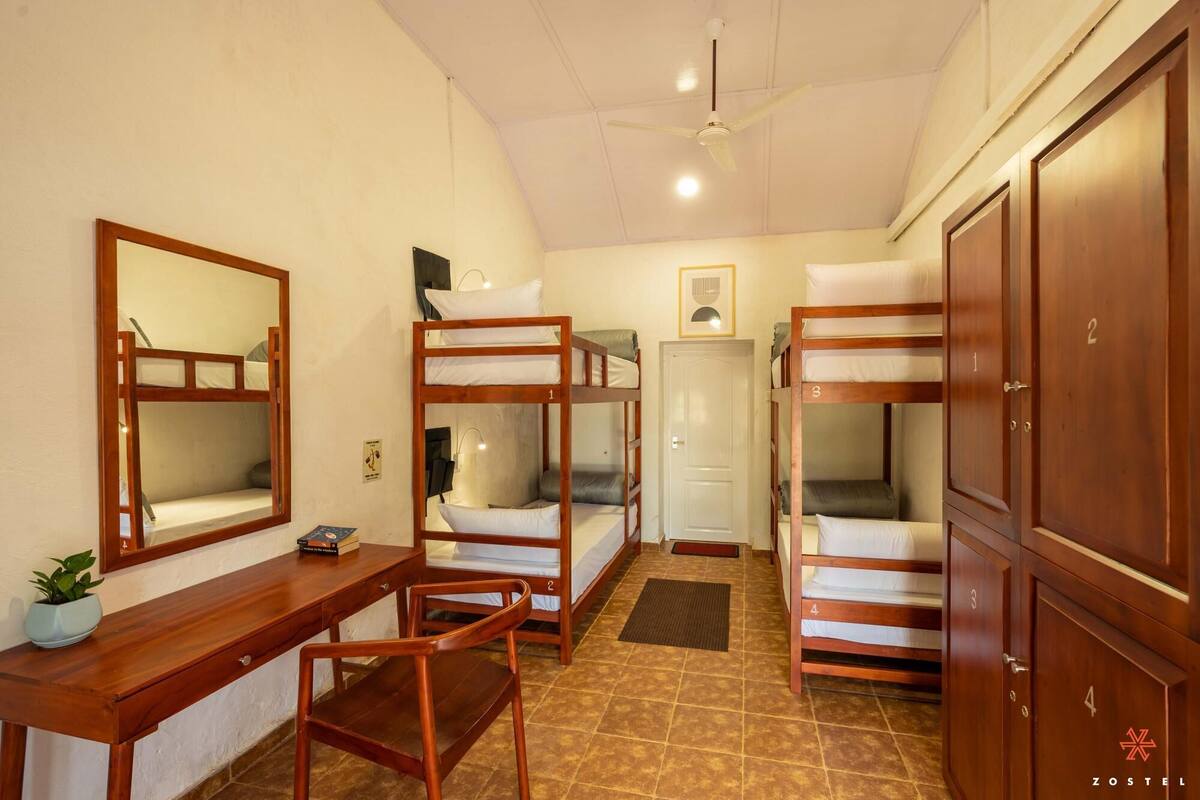 Quarto de dormitório com beliches, uma escrivaninha, uma cadeira, um espelho, um guarda-roupa e um ventilador de teto.