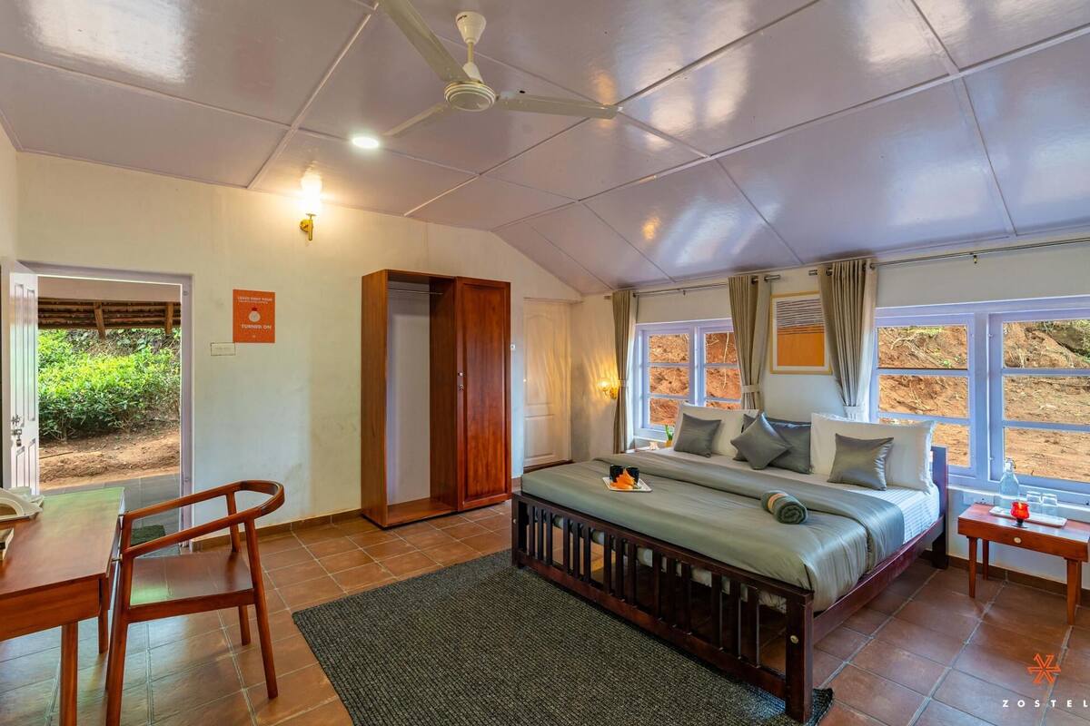 Quarto com cama, escrivaninha, cadeira, janela com cortinas e porta que dá para o exterior.