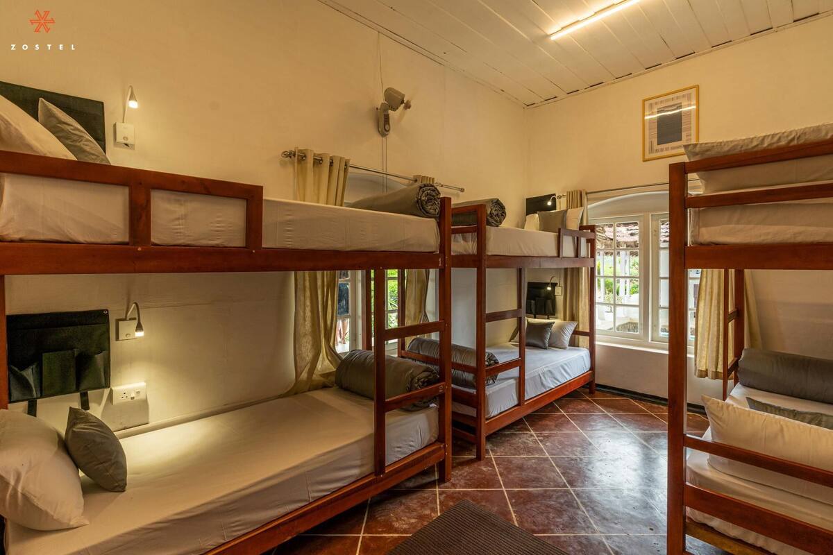 Quarto estilo dormitório com beliches, janela com cortinas e uma pequena escrivaninha com abajur.