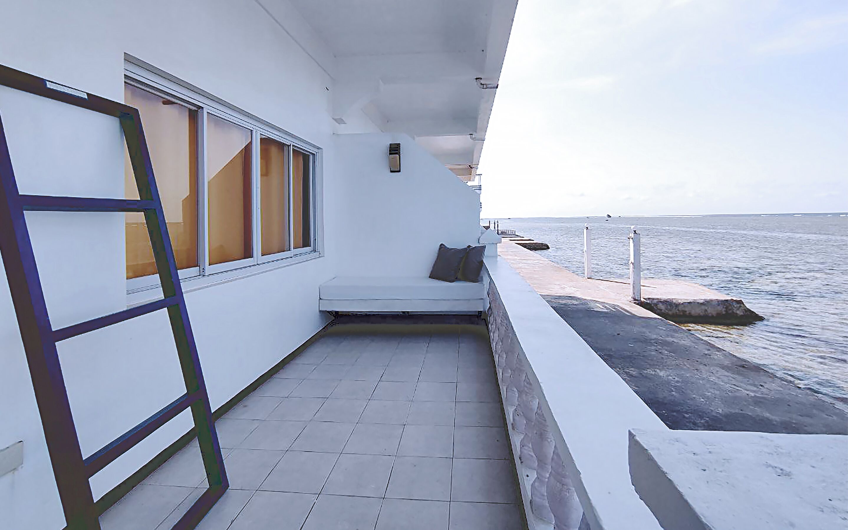 Uma varanda com chão de azulejos brancos, um banco branco com almofadas, uma janela com cortinas laranja e vista para o mar.