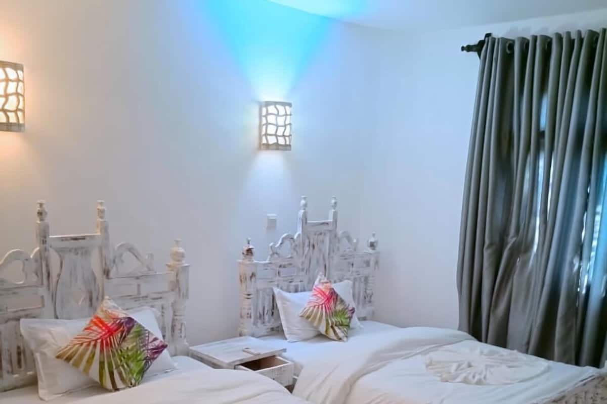Quarto com duas camas, roupa de cama branca, almofadas decorativas, cabeceira e luminária fixada na parede.
