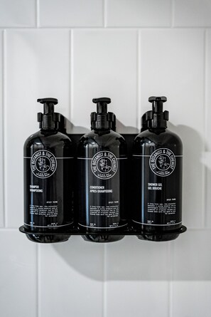 Três frascos pretos de produtos de higiene pessoal da marca The Botanist & The Chemist sobre um revestimento de azulejos brancos.
