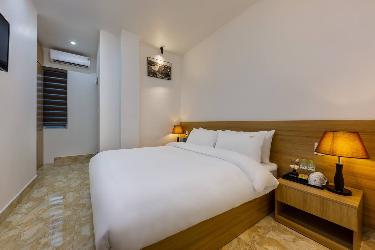 Uma cama bem arrumada com lençóis brancos, cabeceira de madeira e mesa de cabeceira com abajur e bandeja com produtos de higiene.