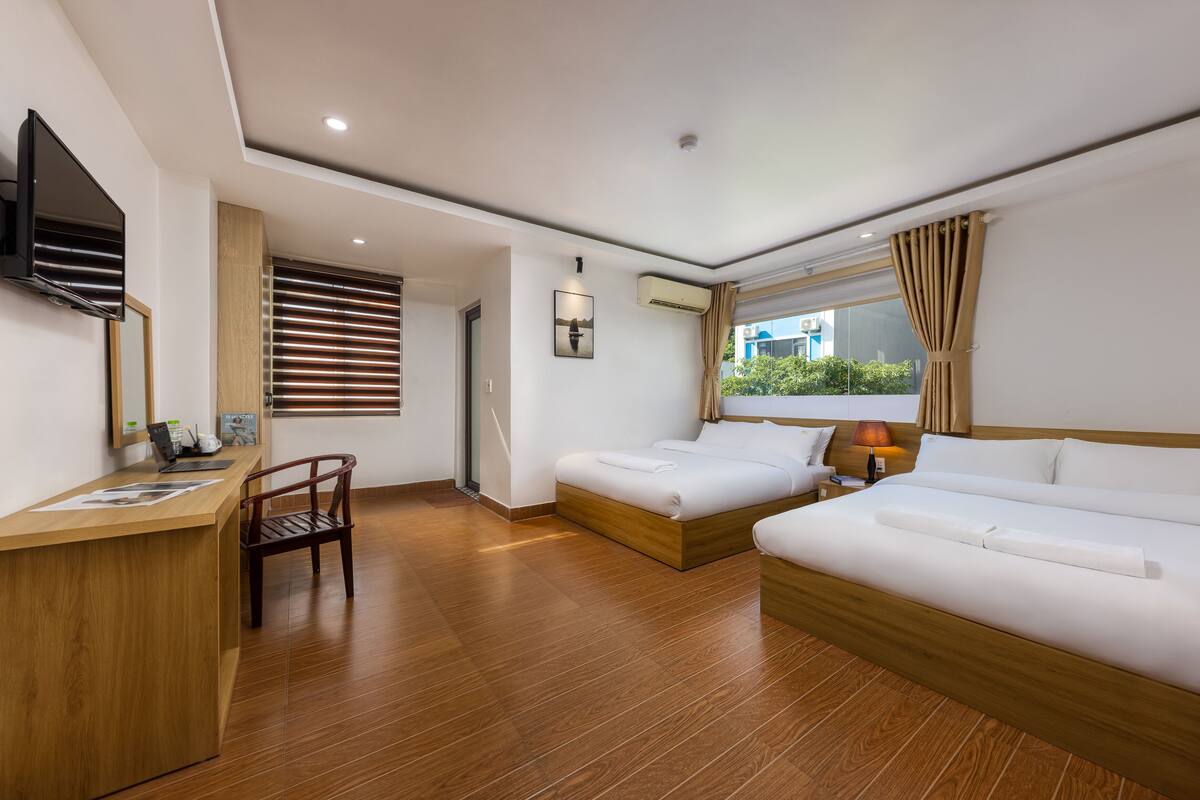 Quarto de hotel com escrivaninha de madeira, duas camas, televisão e janela com cortinas.