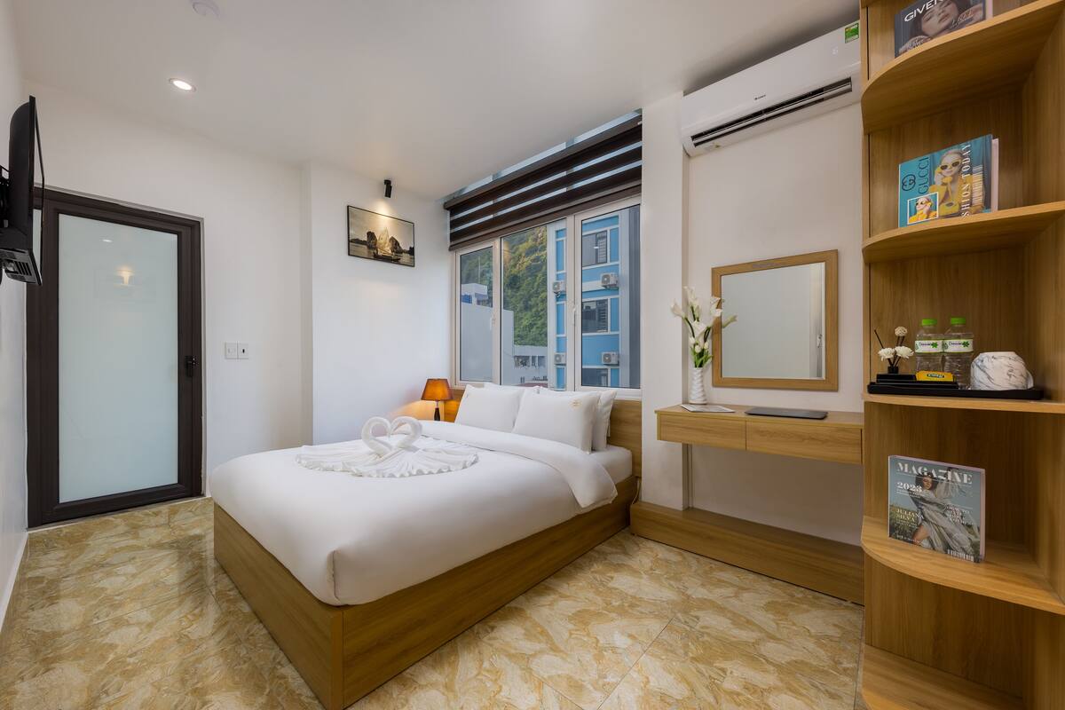Quarto de hotel moderno com uma cama grande, uma mesa de cabeceira de madeira, uma estante e uma janela com vista para edifícios.