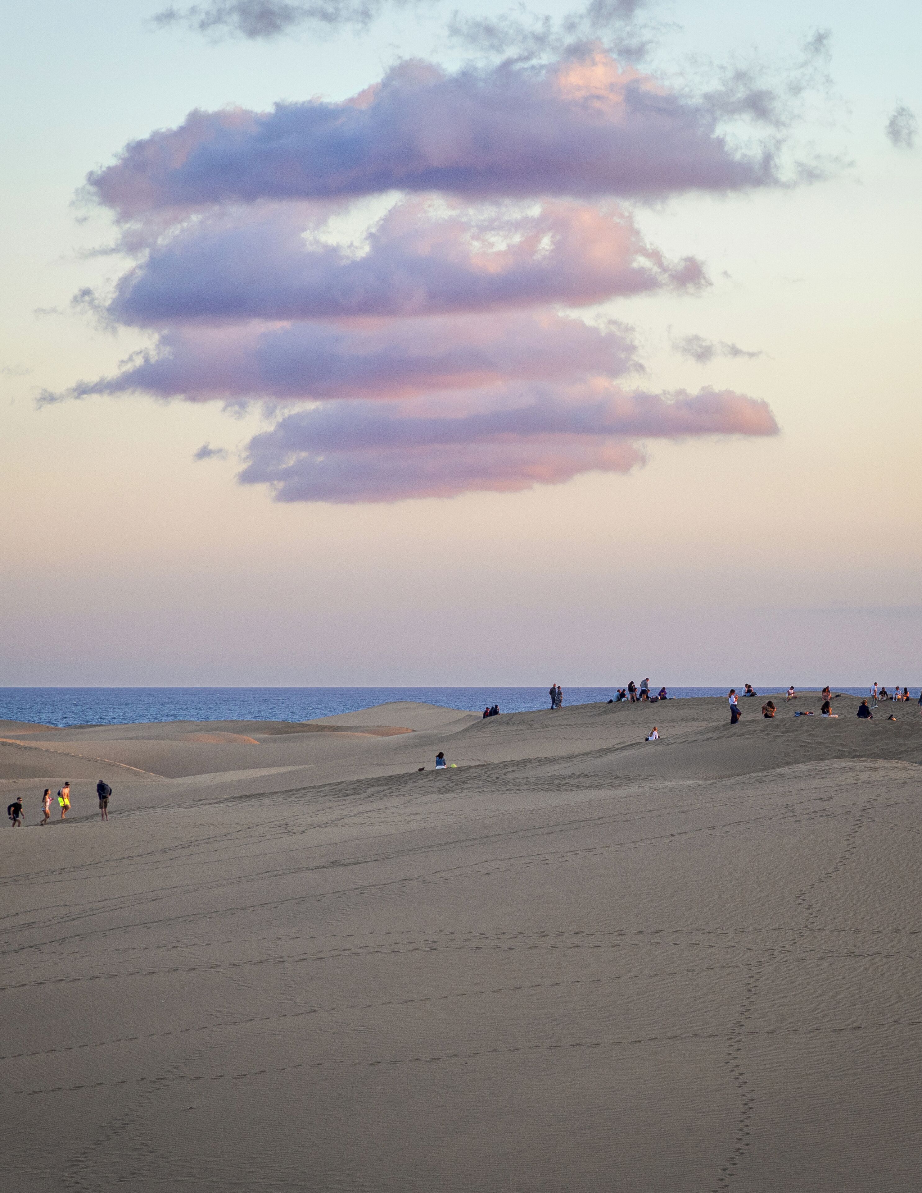 Uma praia com pessoas caminhando e sentadas na areia, e um céu claro com nuvens.