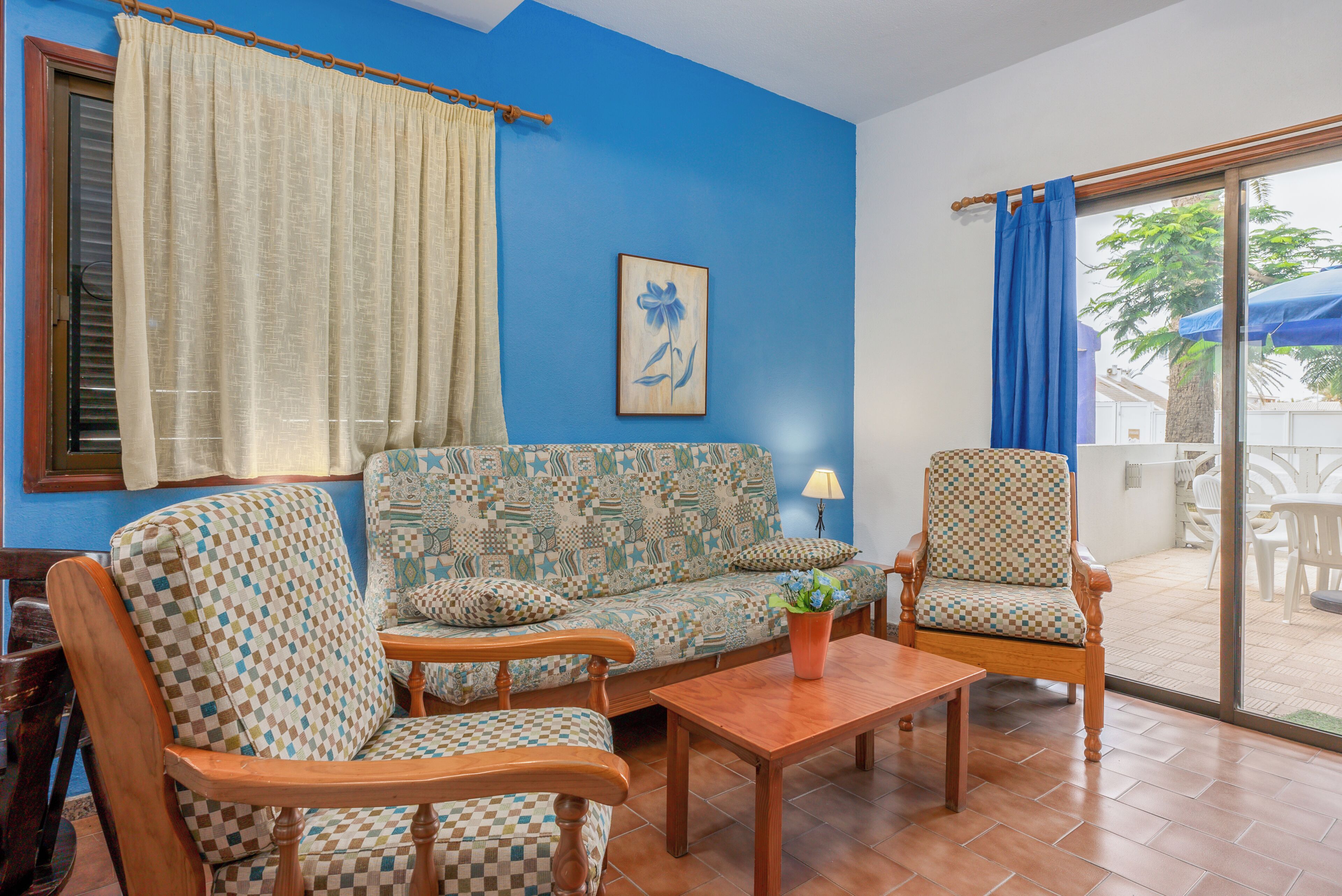 Uma sala de estar com parede azul, sofá estampado, cadeiras de madeira, mesa de madeira e janela com cortinas.