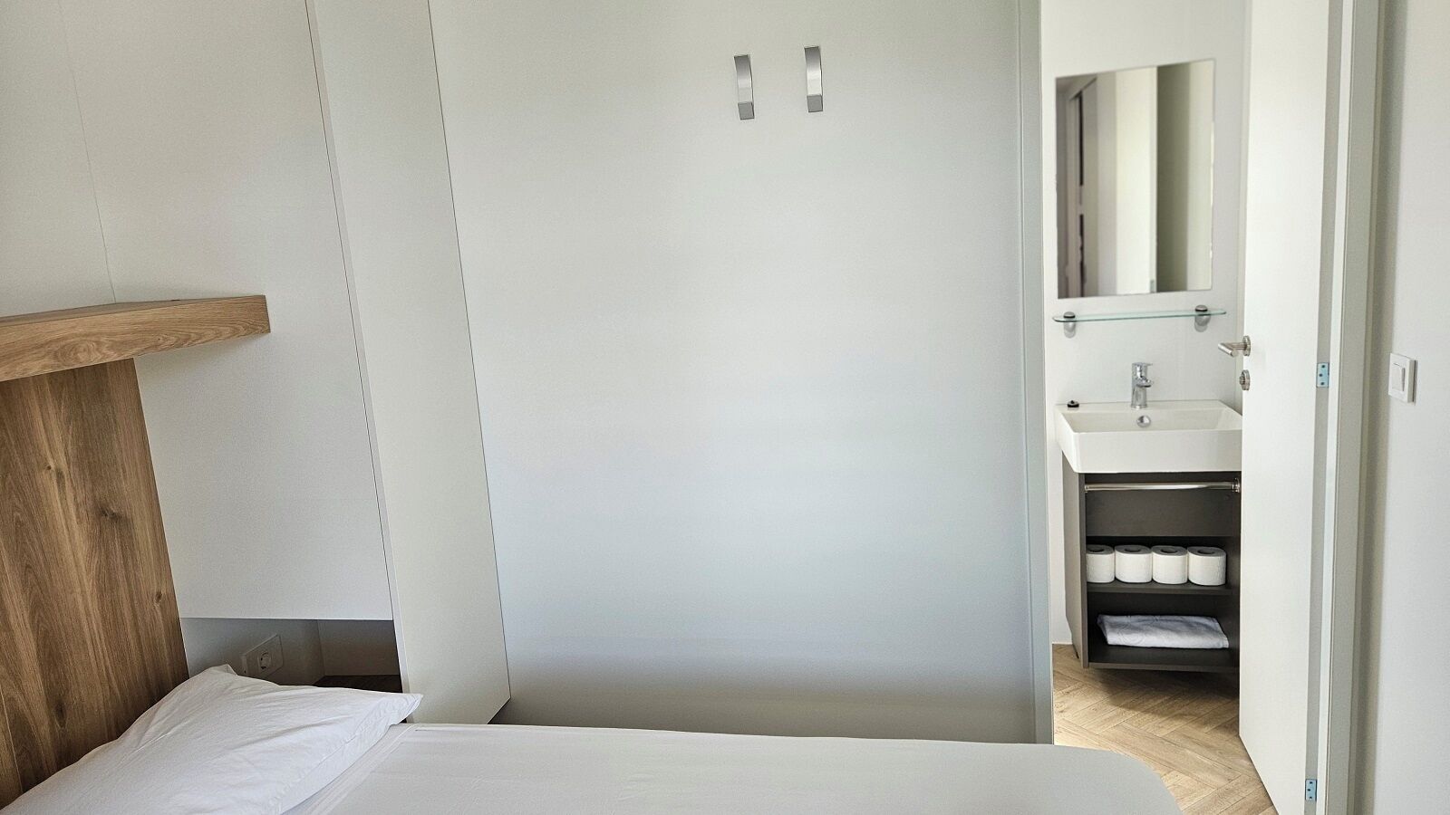 Quarto com cabeceira de madeira, cama branca, guarda-roupa branco e banheiro com espelho, pia e prateleiras.