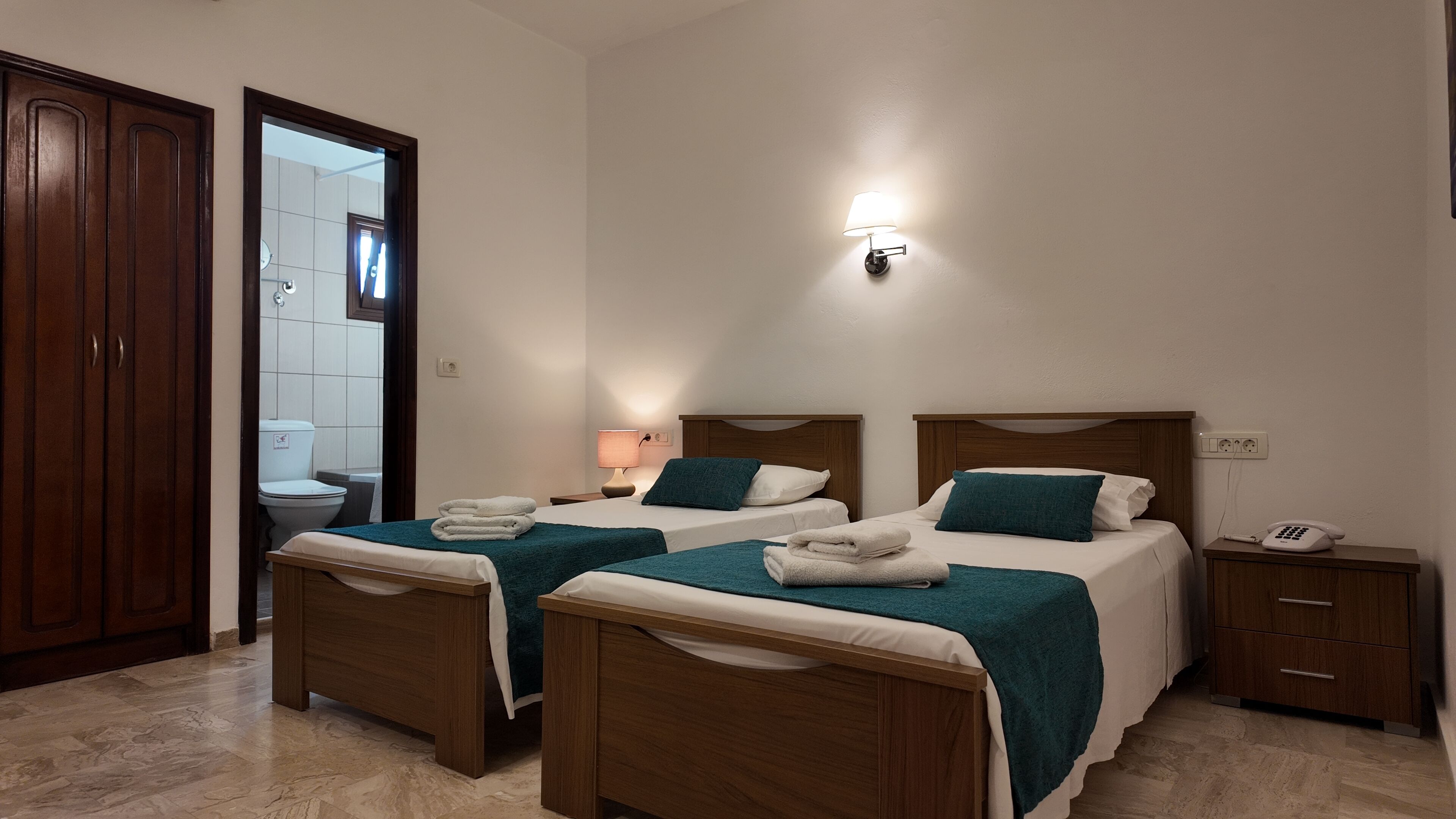 Quarto de hotel com duas camas, um guarda-roupa de madeira, uma mesa de cabeceira com abajur e um telefone.