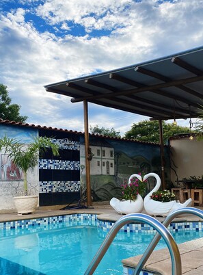 Uma piscina com dois vasos em forma de cisne, um mural de azulejos e uma área de estar coberta.