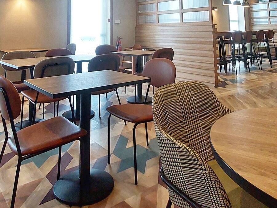 Uma sala de jantar moderna com várias mesas e cadeiras, um piso estampado e uma janela grande que permite entrada de luz natural.