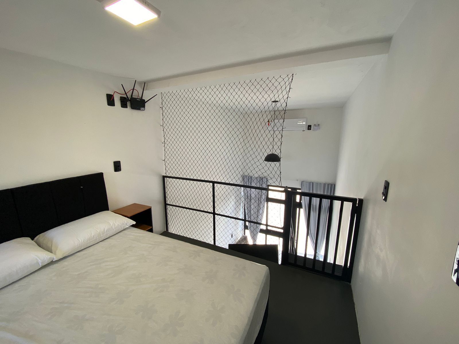 Quarto com cama, mesa de cabeceira, escrivaninha, cadeira, janela com cortinas e varanda com guarda-corpo.