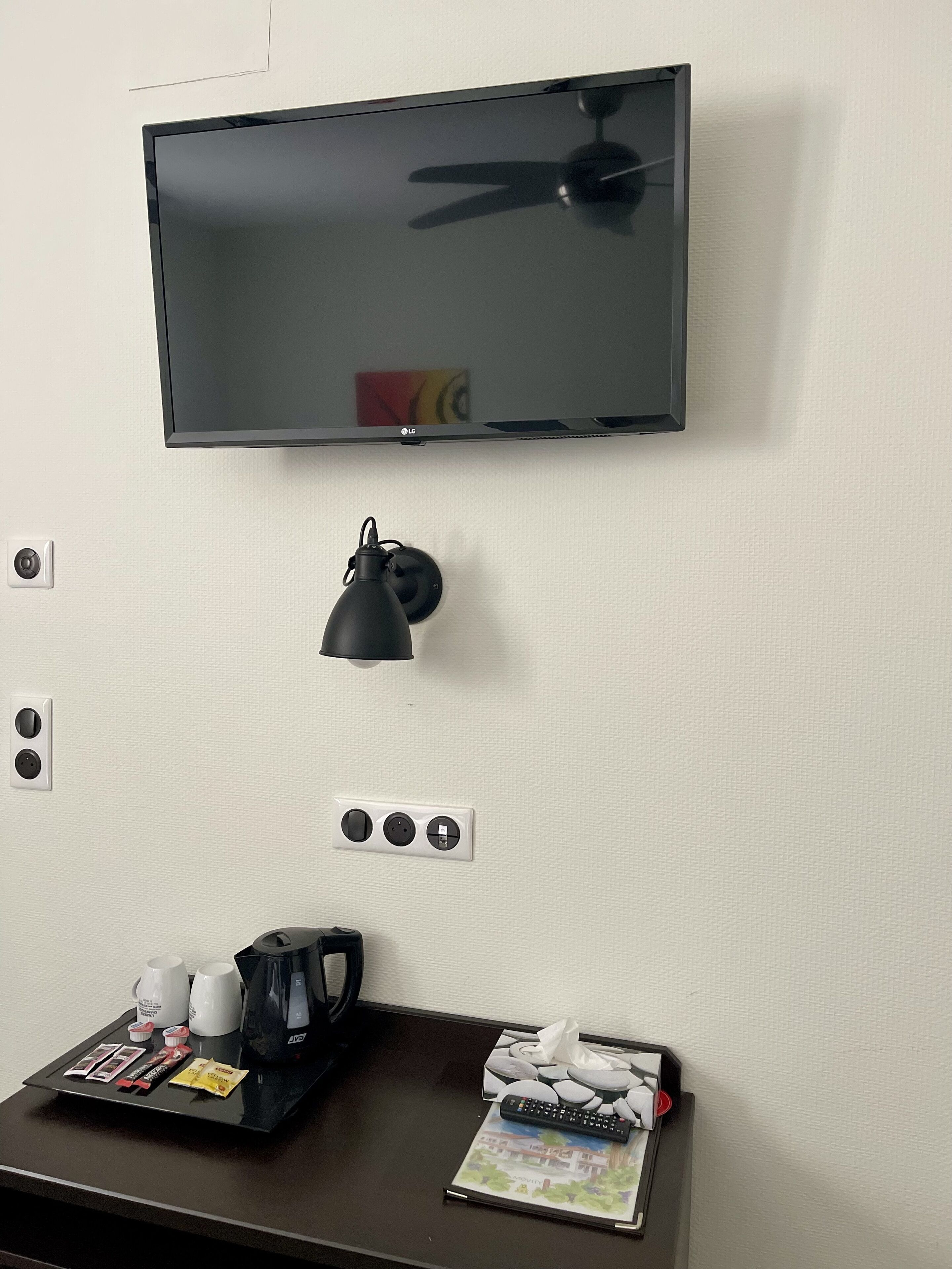 Uma TV de tela plana fixada na parede, um abajur preto, uma tomada, e uma mesa com um controle remoto, lenços de papel e uma revista.