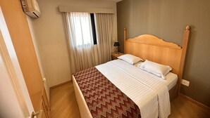 Quarto de hotel com cama de madeira, janela com cortinas, abajur e ar condicionado fixado na parede.