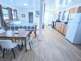 Quarto compacto com cama, mesa de jantar com cadeiras, cozinha equipada com geladeira e janela com cortinas.