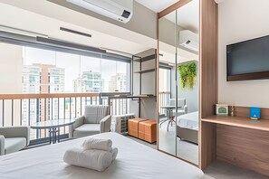 Quarto de hotel moderno com uma cama grande, uma escrivaninha, uma cadeira, uma mesa pequena, uma TV e uma varanda com vista para a cidade.