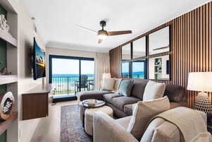 Uma sala de estar moderna com sofá, banquinho e ventilador de teto. Há uma janela grande com vista para o oceano.